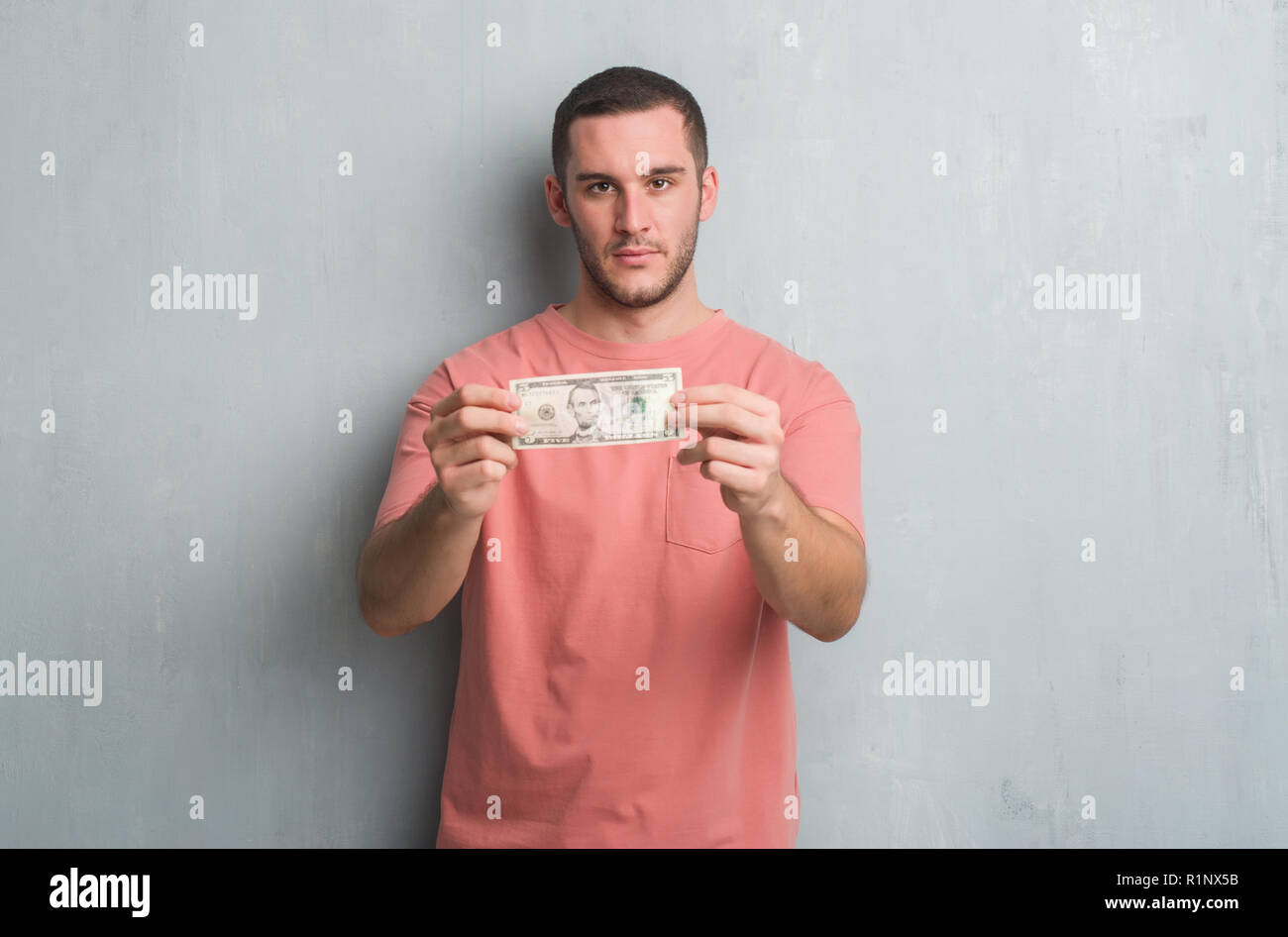 Young caucasian man over grey grunge wall showing five dollars with a ...