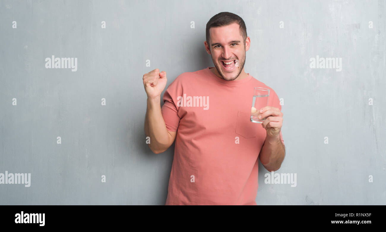 Young caucasian man over grey grunge wall drinking a glass of water ...