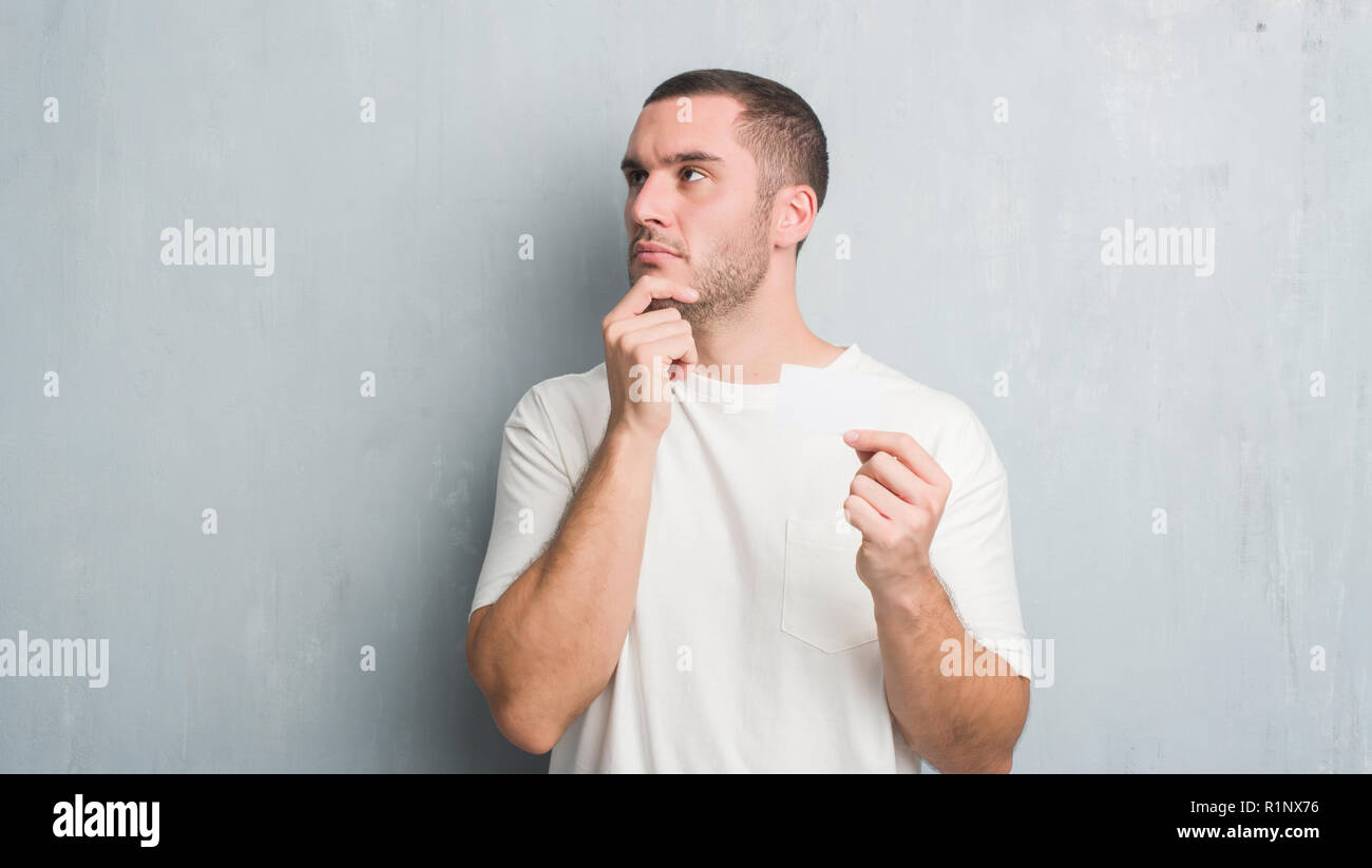 Young caucasian man over grey grunge wall showing blank visit card ...