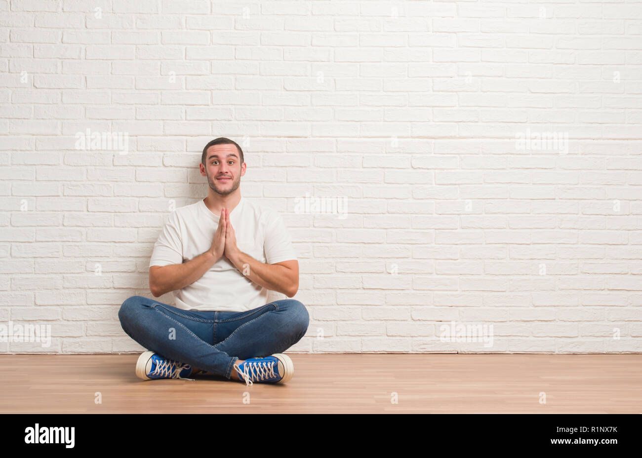 Young caucasian man sitting on the floor over white brick wall praying ...