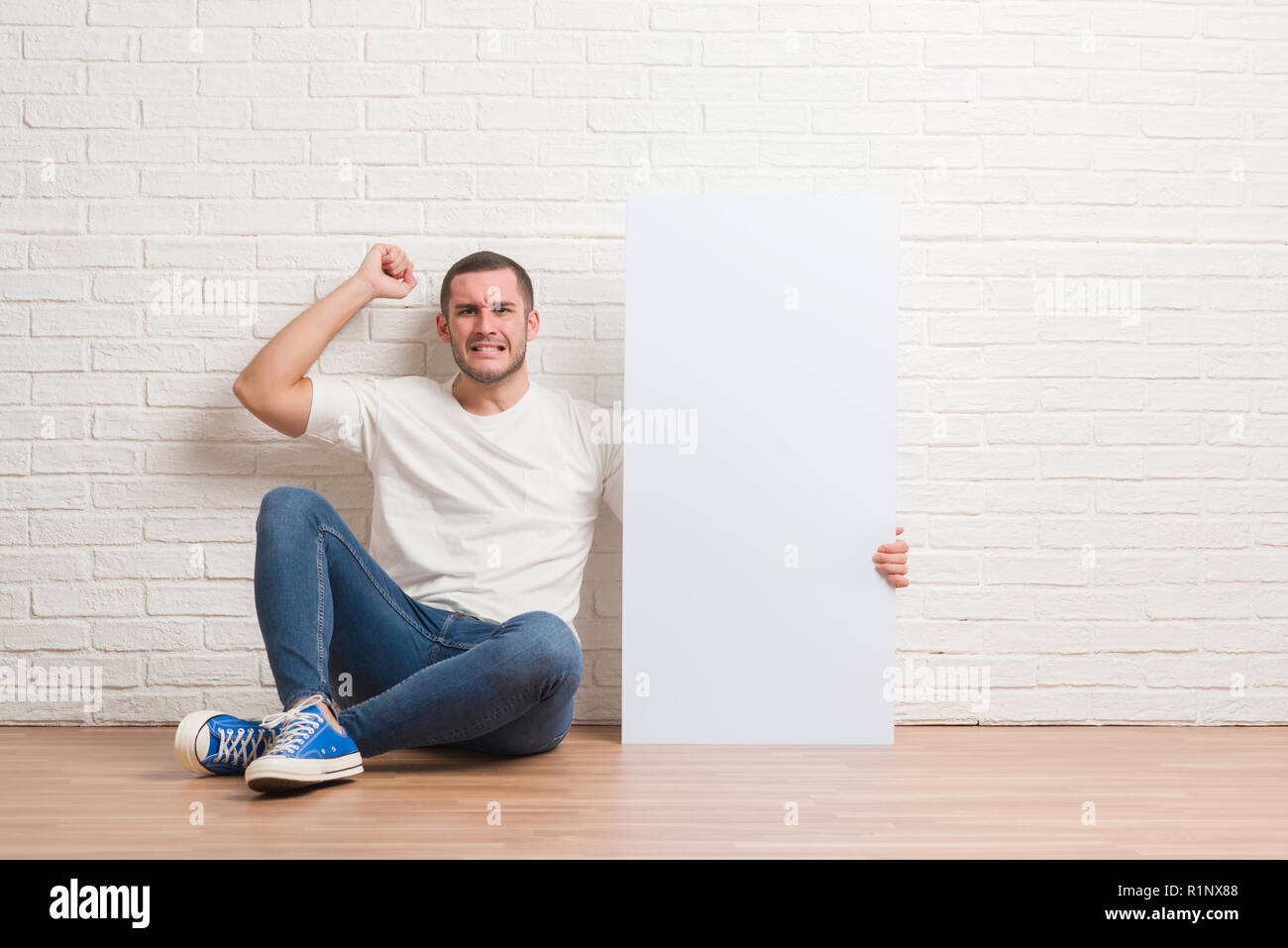 Young caucasian man sitting over white brick wall holding bottle of ...