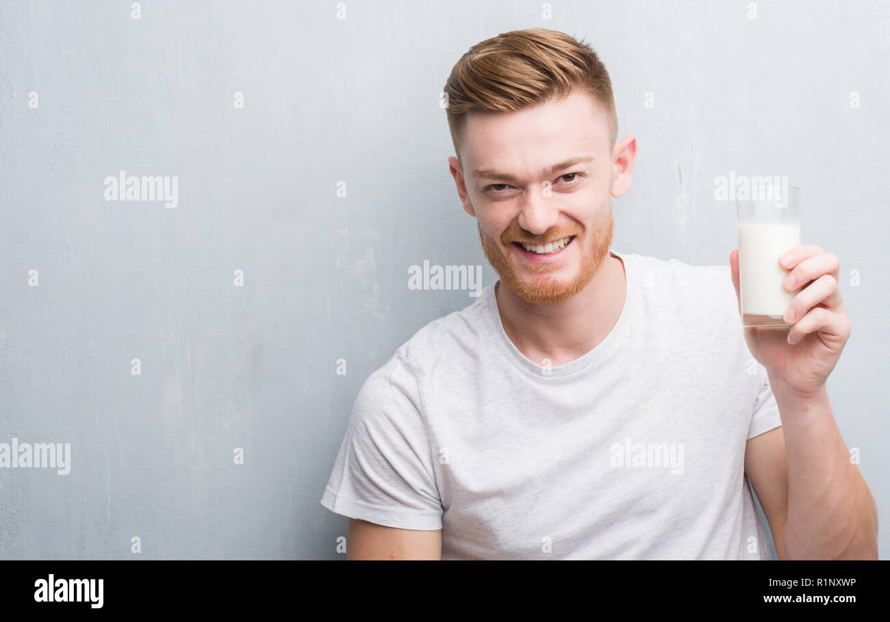 Young redhead man over grey grunge wall drinking a glass of milk with a ...
