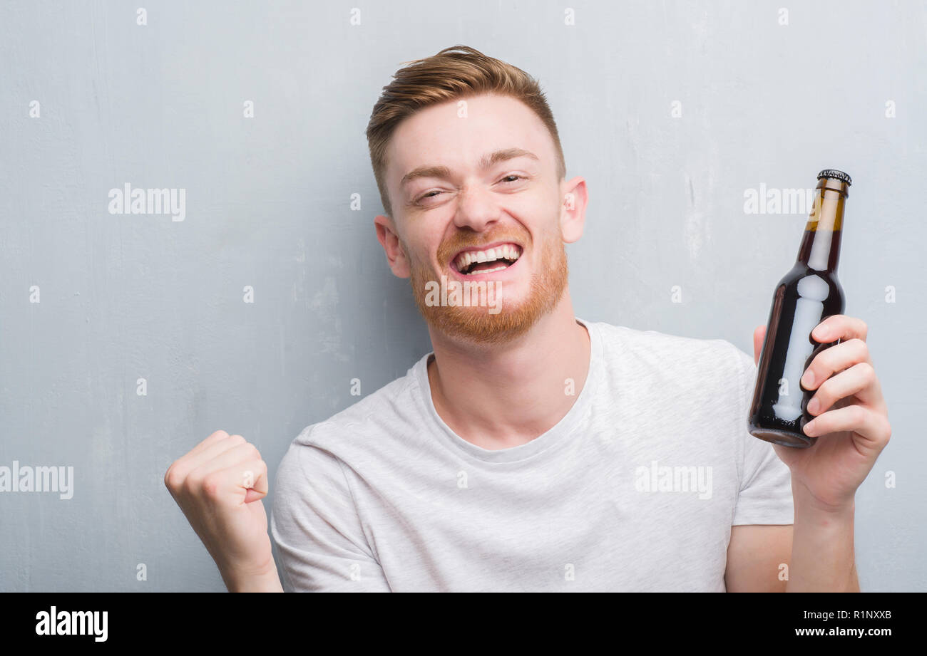 Young redhead man over grey grunge wall drinking beer bottle screaming ...
