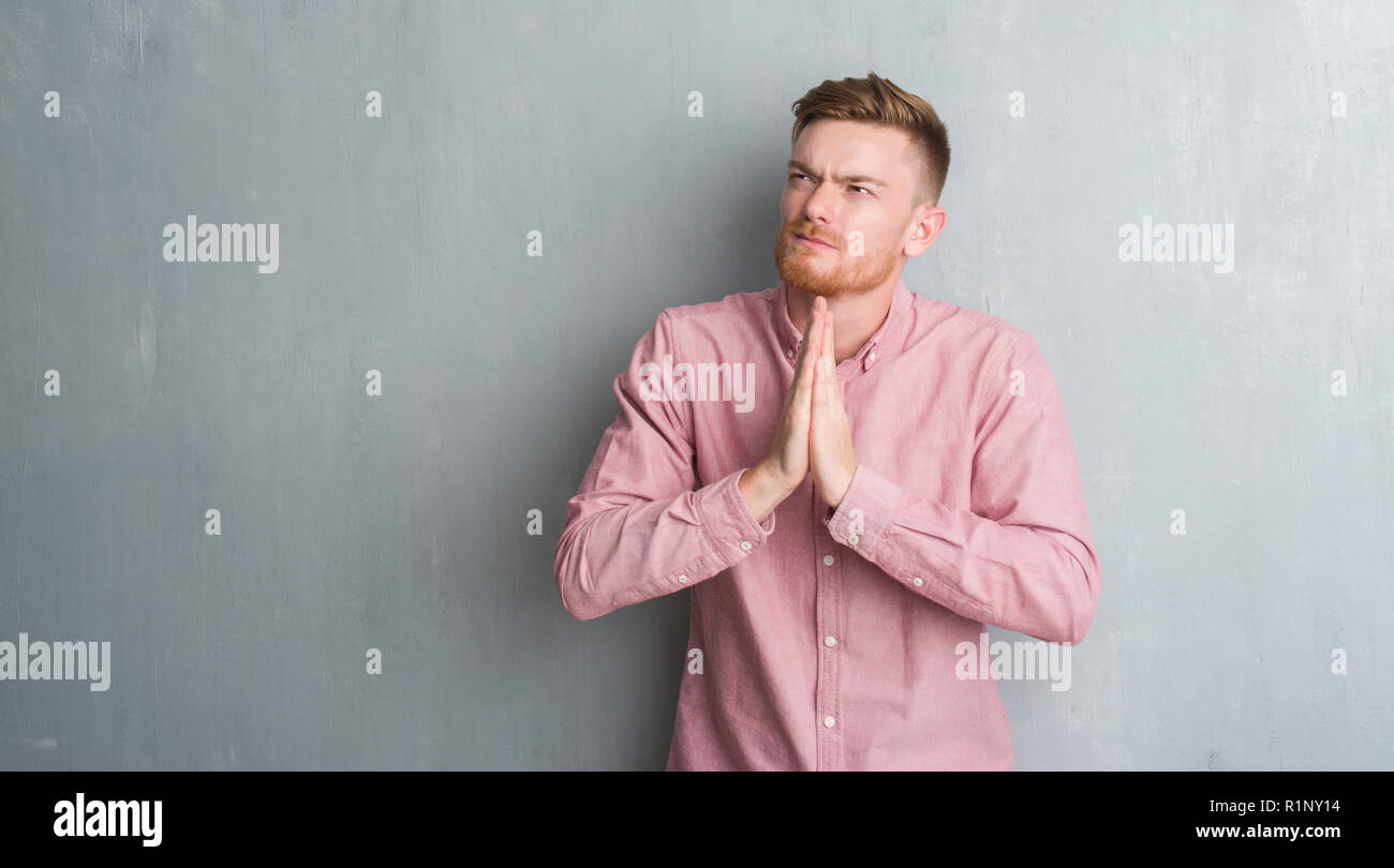 Young redhead man over grey grunge wall wearing pink shirt begging and ...