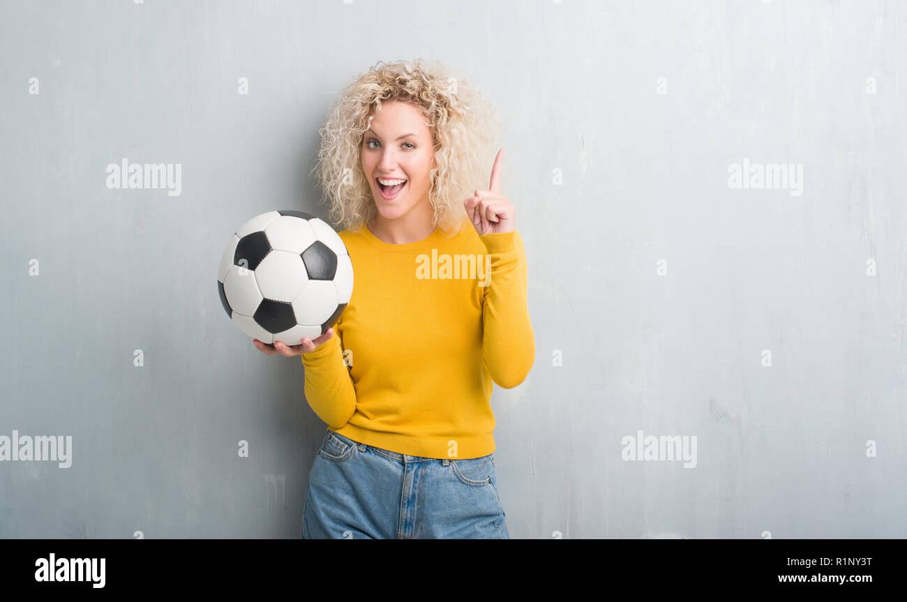 Young blonde woman over grunge grey background holding soccer football ...
