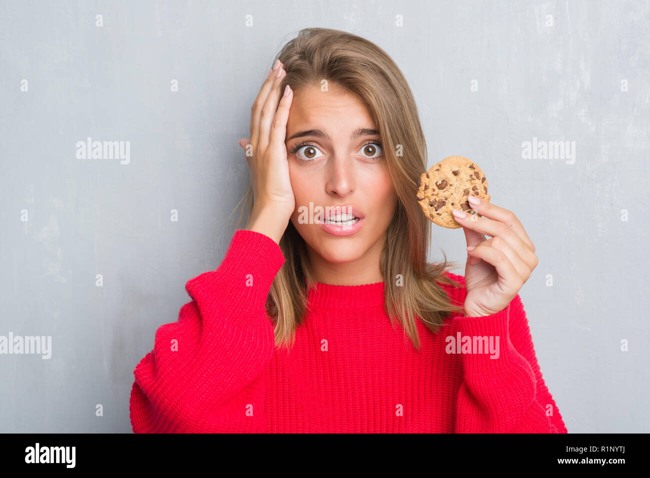Beautiful young woman over grunge grey wall eating chocolate chip cooky ...