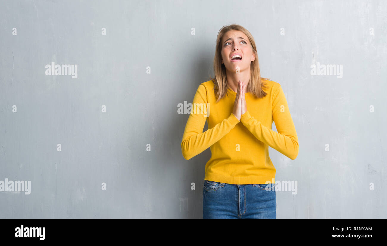 Beautiful young woman standing over grunge grey wall begging and ...