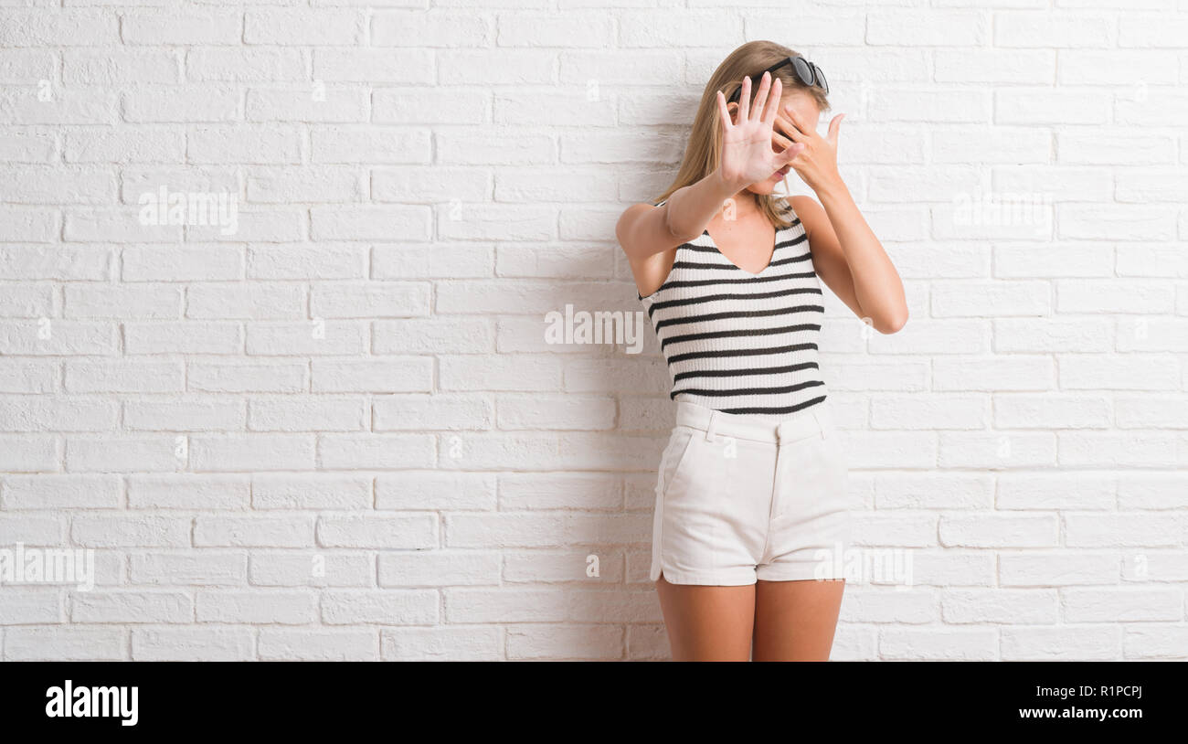 Young beautiful blonde woman over white brick wall covering eyes with ...