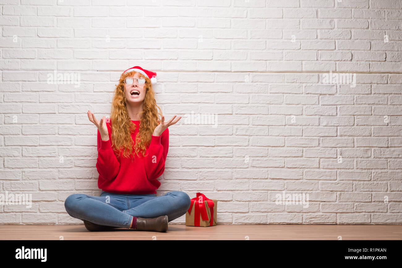 Young redhead woman sitting over brick wall wearing christmas hat crazy ...