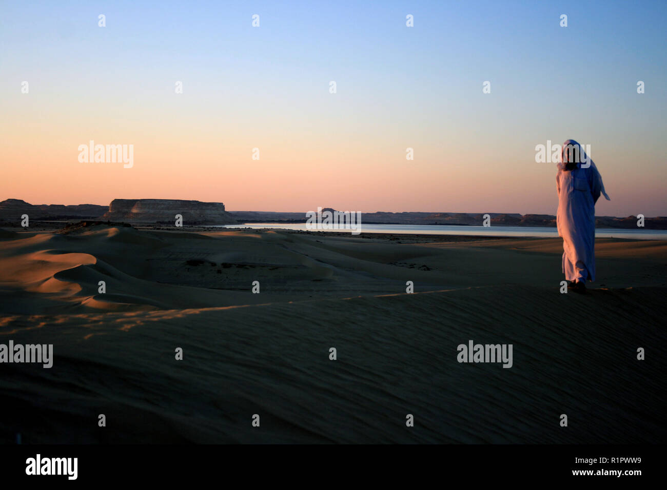 Side-lit man in long robe walking along sand dunes under sunset in the Sahara Desert near Siwa ...