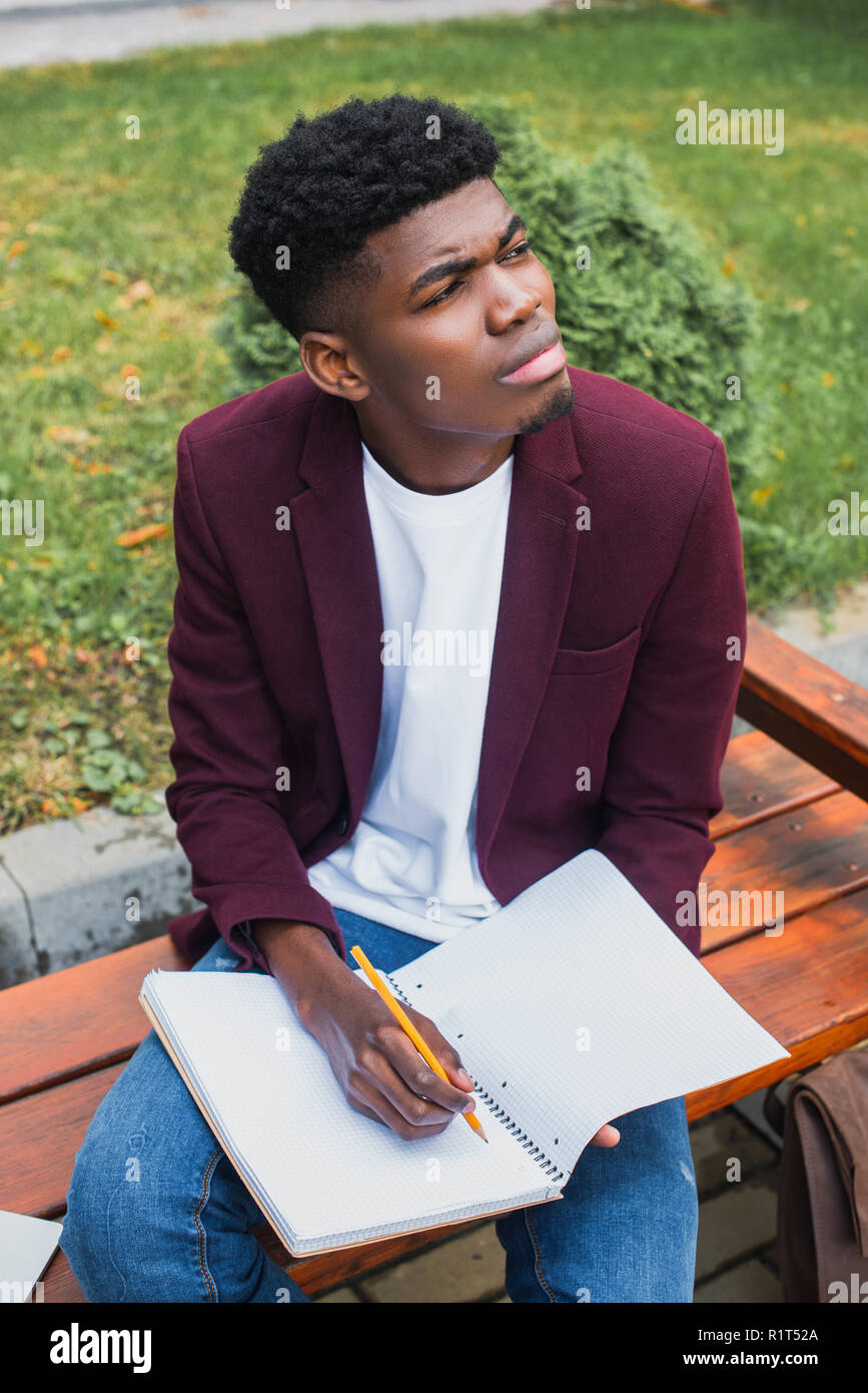thoughtful young man sitting on bench and writing in blank notebook ...