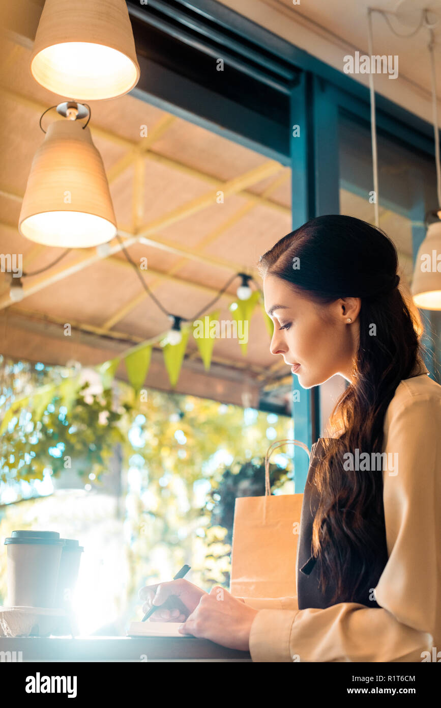 side view of attractive waitress in apron writing something to notebook ...