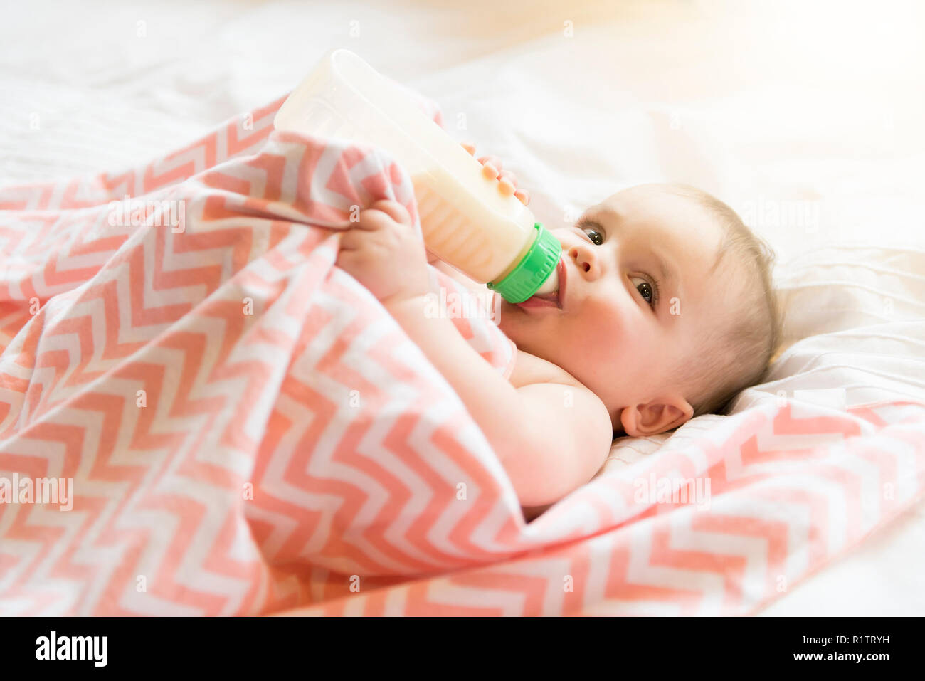 A Little child girl lying on white bed and drinking milk from bottle ...