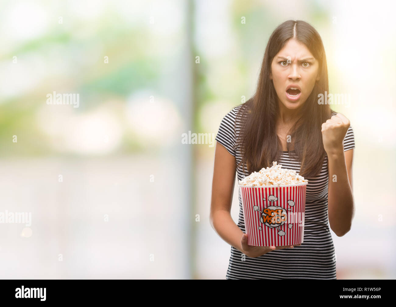 Young beautiful hispanic eating popcorn annoyed and frustrated shouting ...