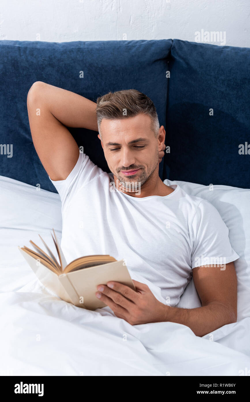 high angle view of man reading book while laying in bed during morning ...