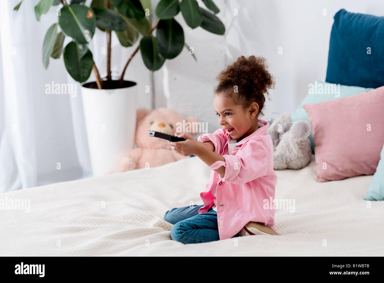 Emotional African american child sitting on the bed with tv remote and ...