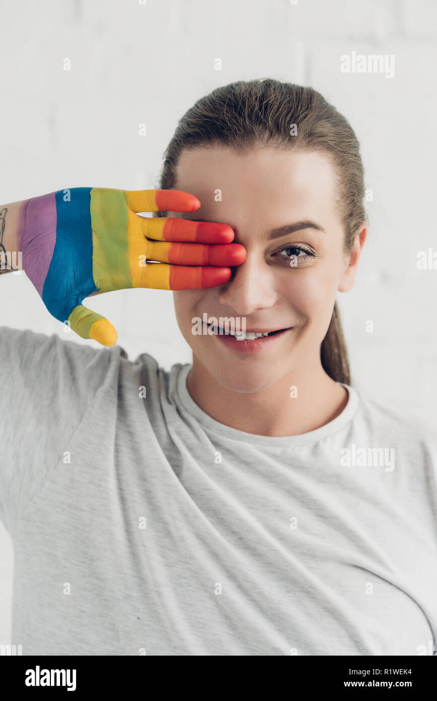 smiling transgender man covering eye with hand painted in colors of ...