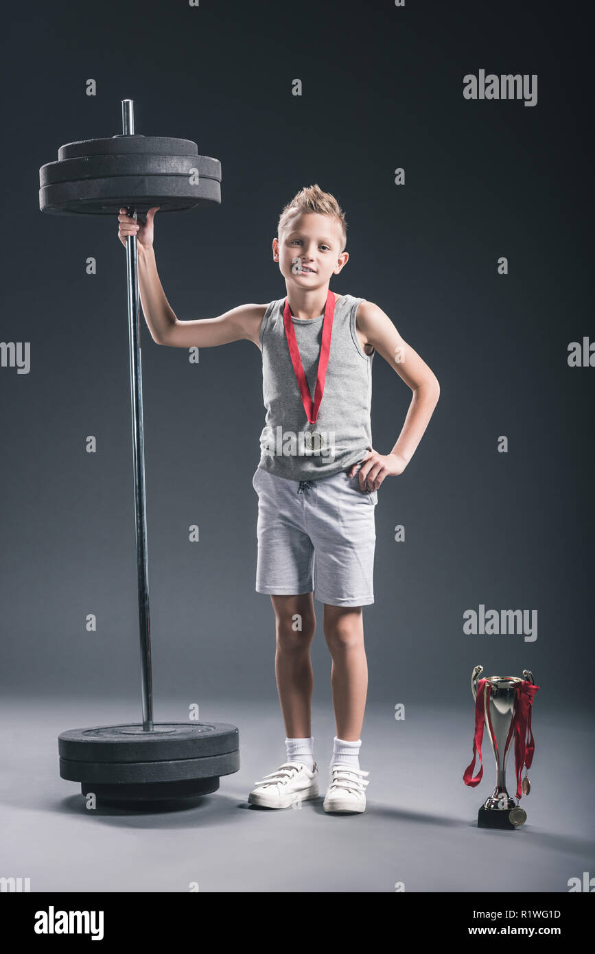 pre-adolescent boy in sportswear with barbell, champions cup and medals ...