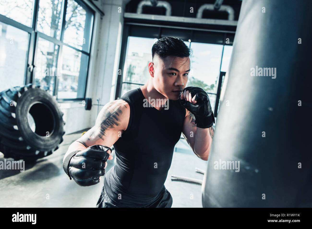 young sportsman wearing boxing gloves and punching boxing bag at gym ...