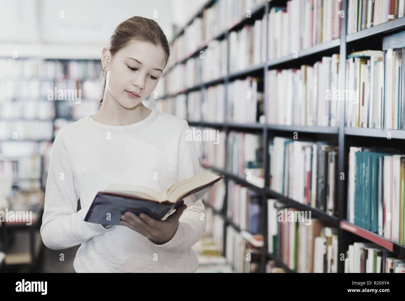 Intelligent cheerful positive preteen girl standing alone near bookcase ...