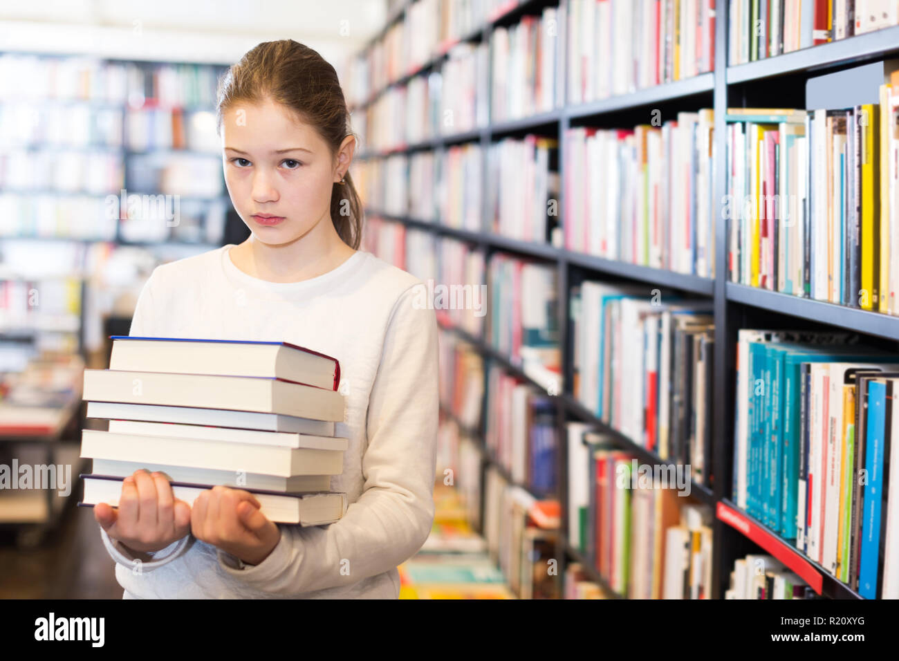 Sad tired tweenager standing in library with pile of books in hands ...