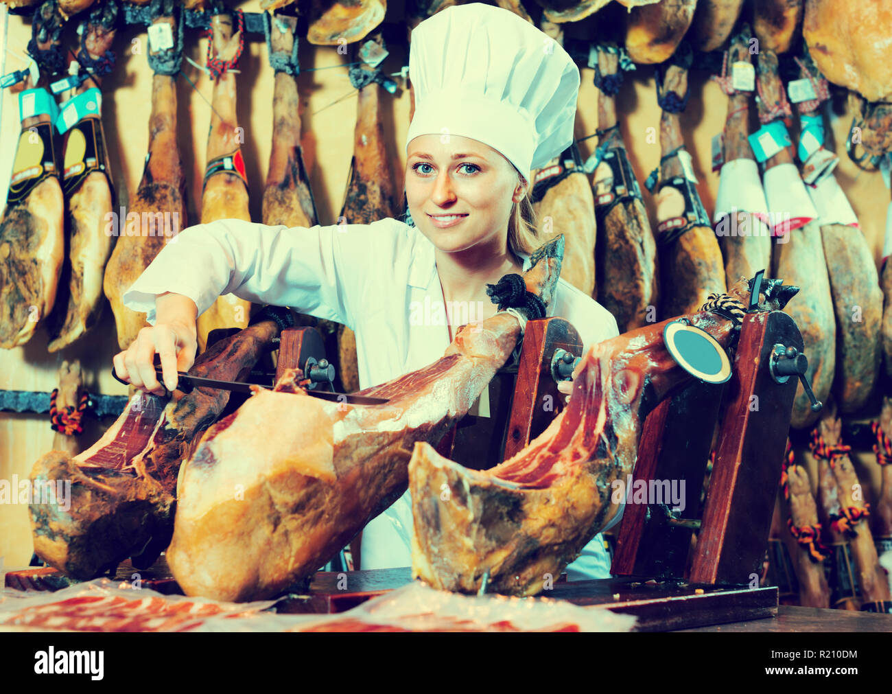Portrait of young glad woman cutting dried ham in delicatessen meat ...