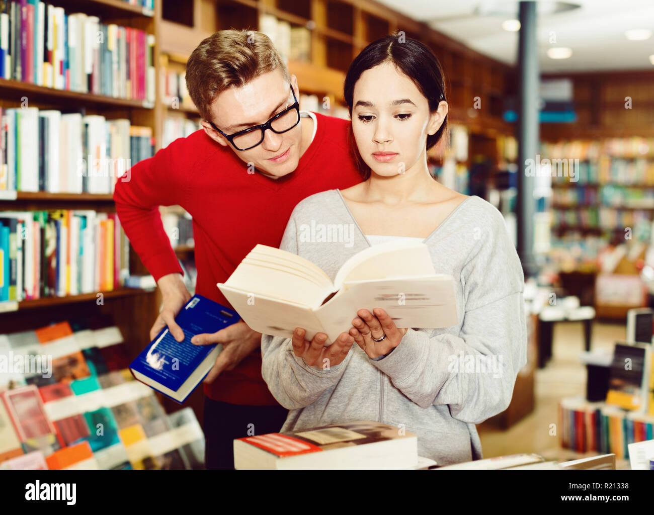 Cheerful positive smiling girl reading book in bookstore while guy ...