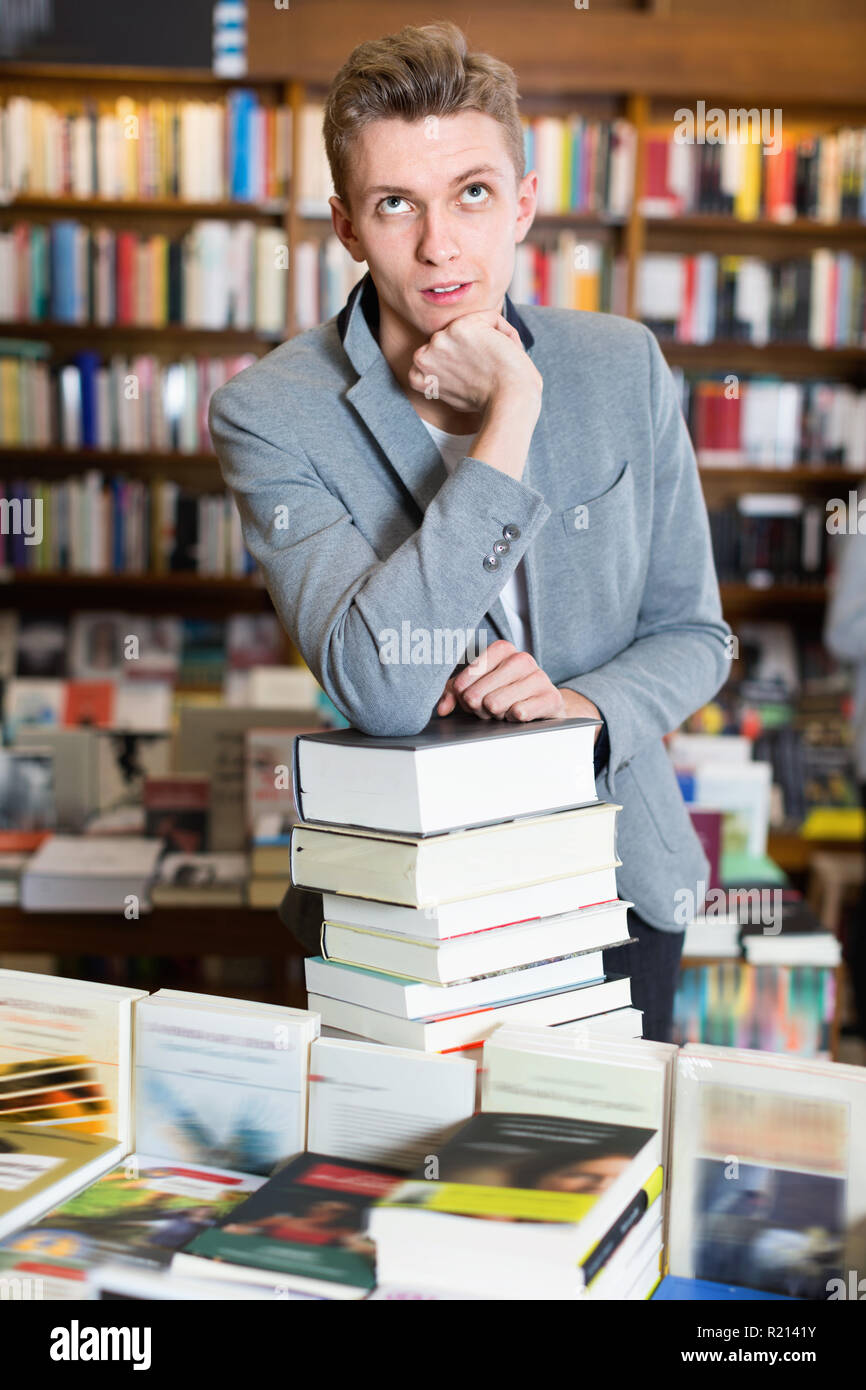Pensive cheerful positive young man posing on stack of books in ...