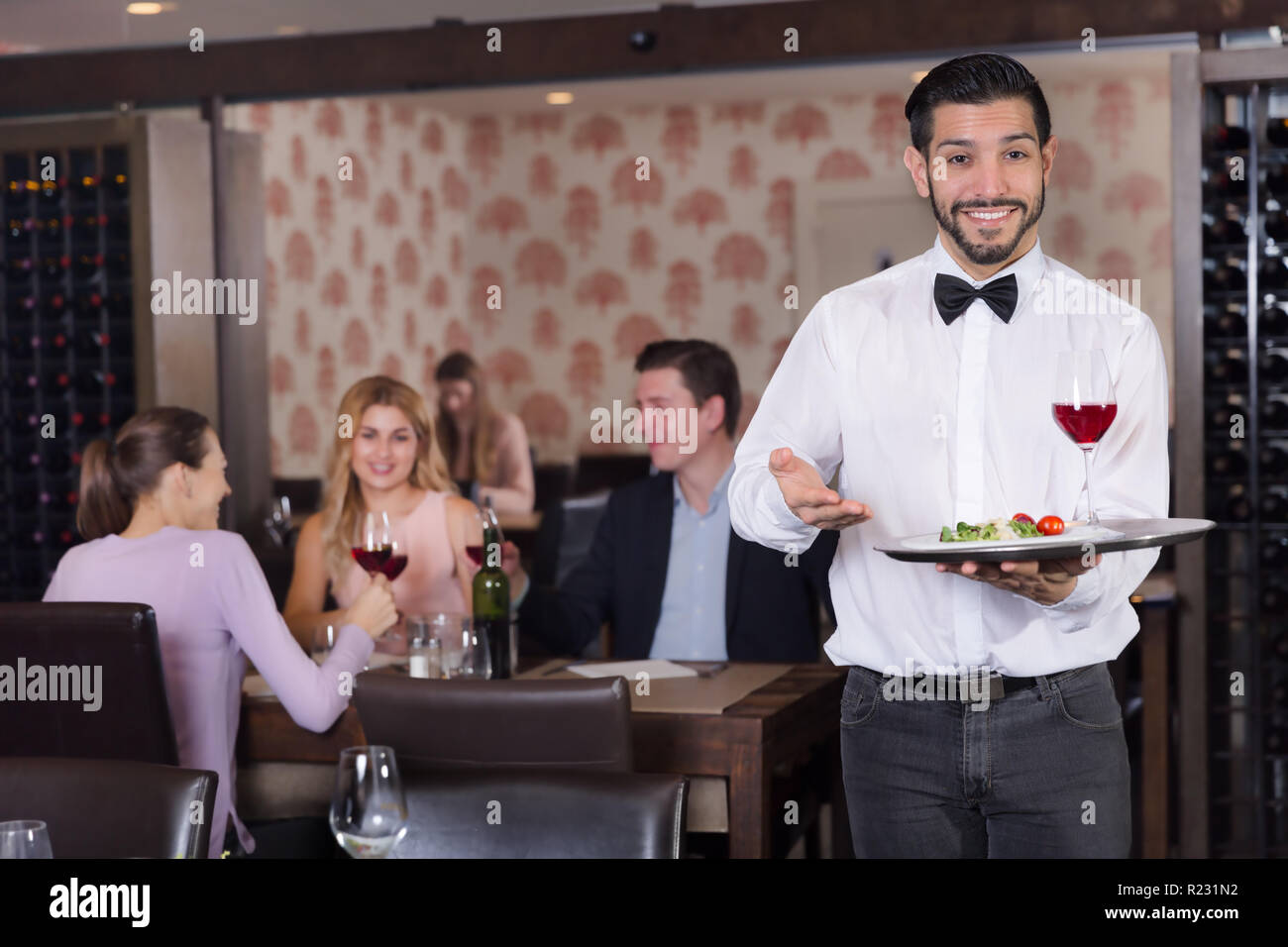 Polite waiter holding tray at a restaurant with customers his behind ...
