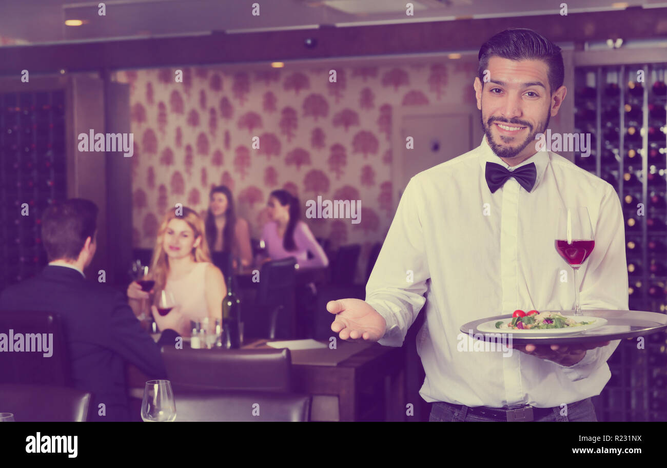Polite waiter holding tray in a restaurant with customers his behind ...