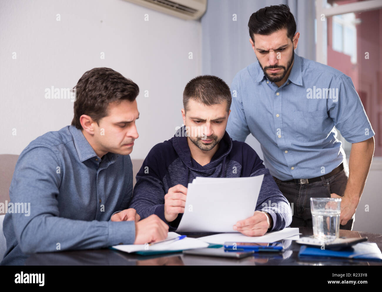 Three upset male friends looking worriedly at papers at home table ...