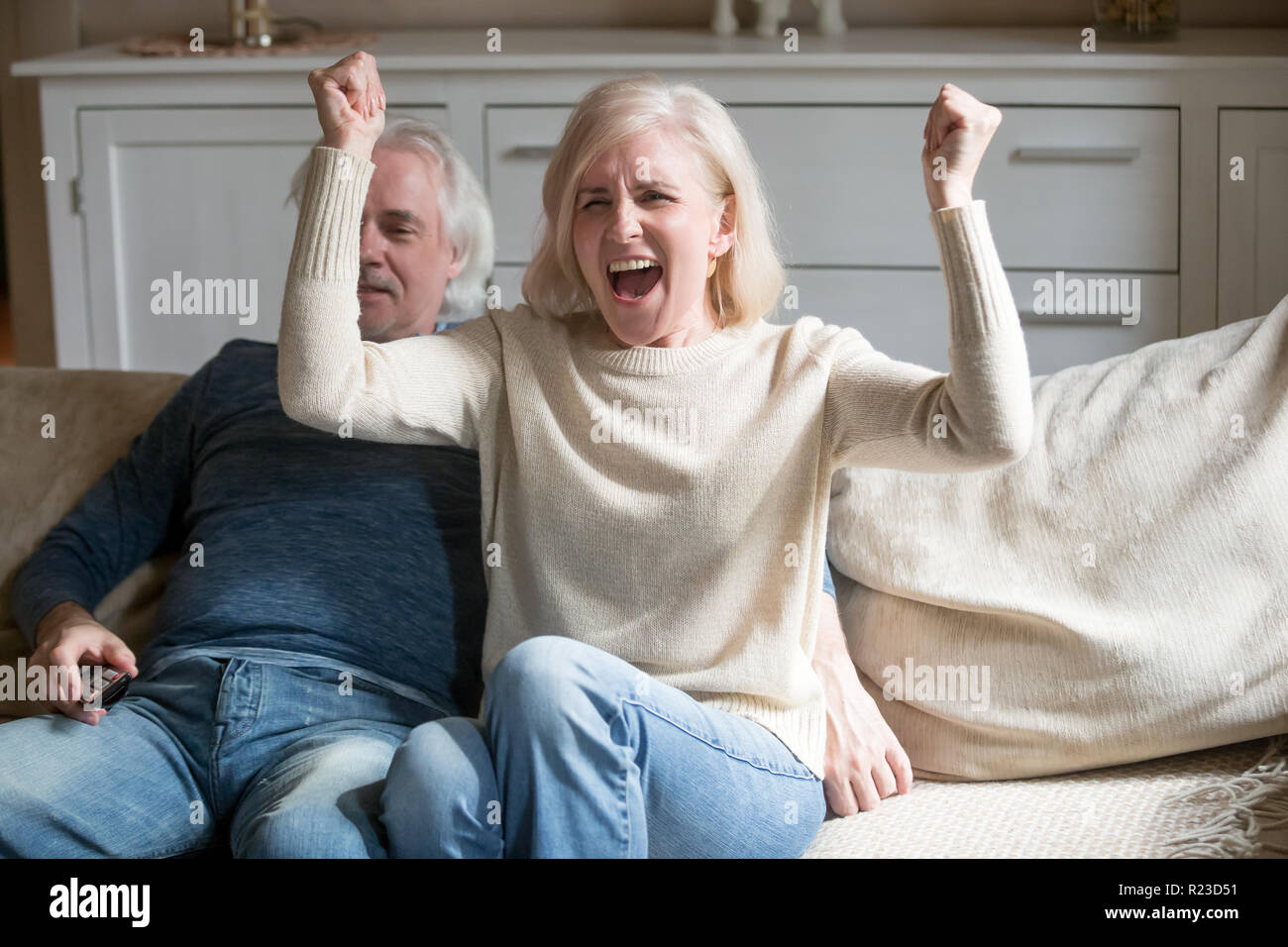 Happy aged couple relax on cozy couch at home watching football game on TV, excited senior wife ...