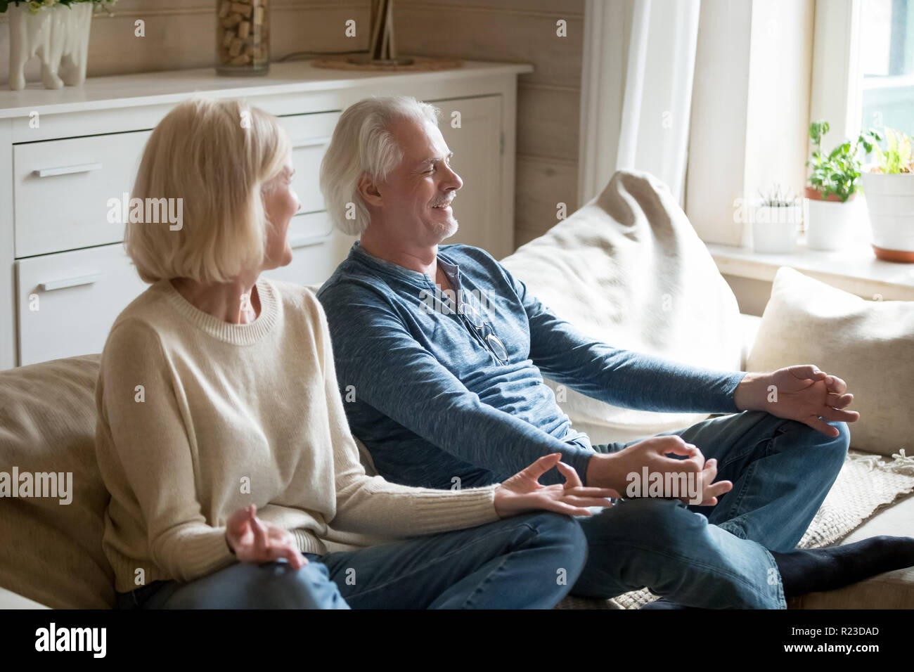 Smiling aged husband and wife sit on couch meditating in lotus position ...