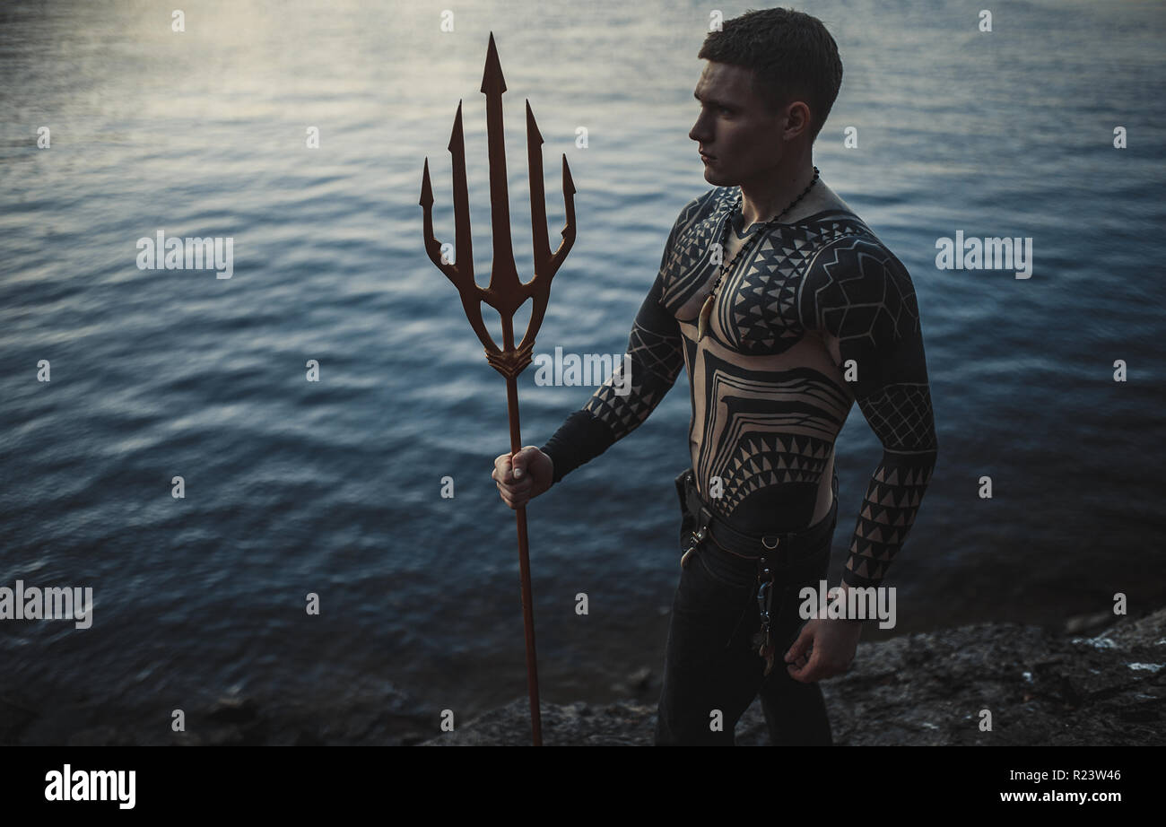A young man in armor with a trident in his hands against the background ...