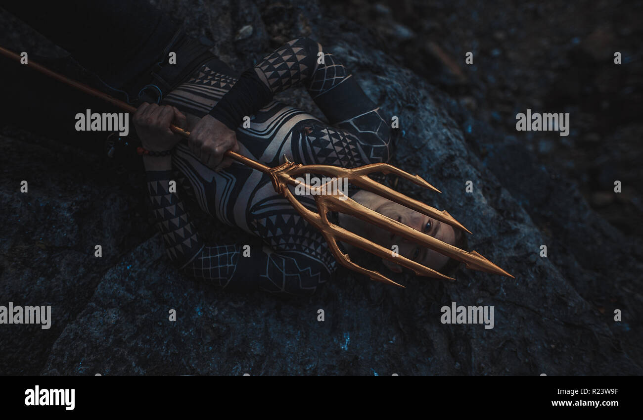 A young man in armor and with a trident in his hands lies on the rock ...