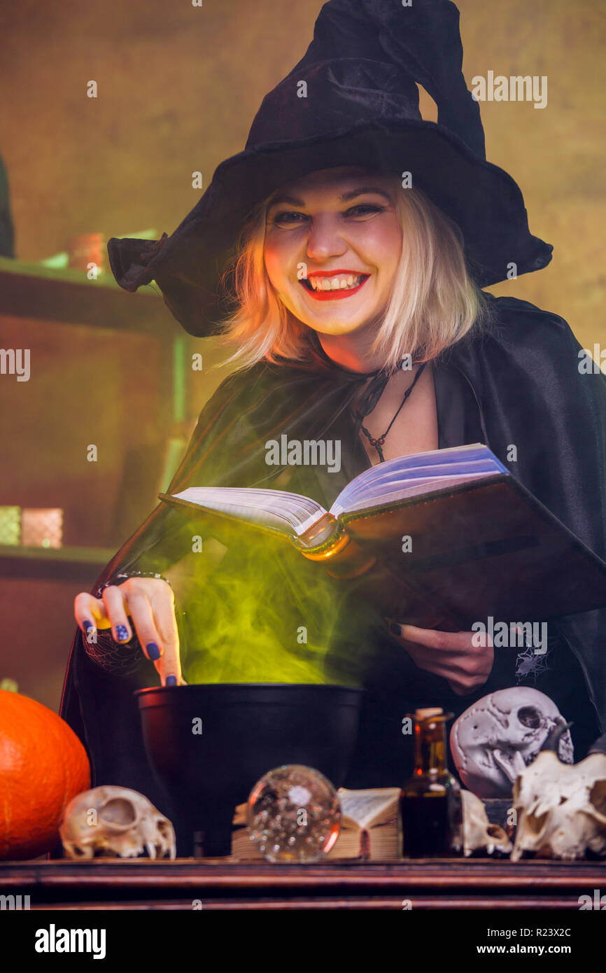 Photo of smiling witch blonde in black hat with book potting potion in ...