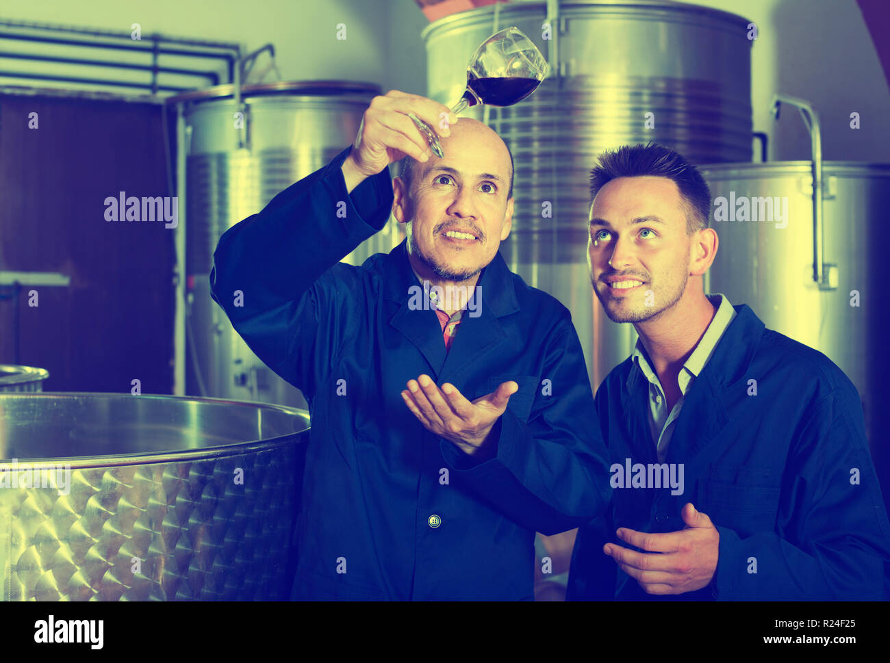 two smiling winery male workers in uniform looking at wine sample in ...