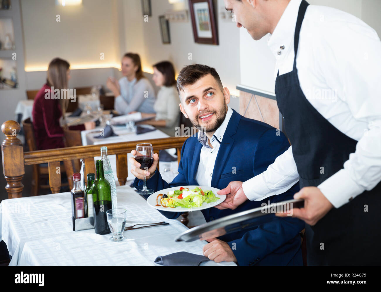 Glad positive smiling waiter serving delicious salads to handsome young ...