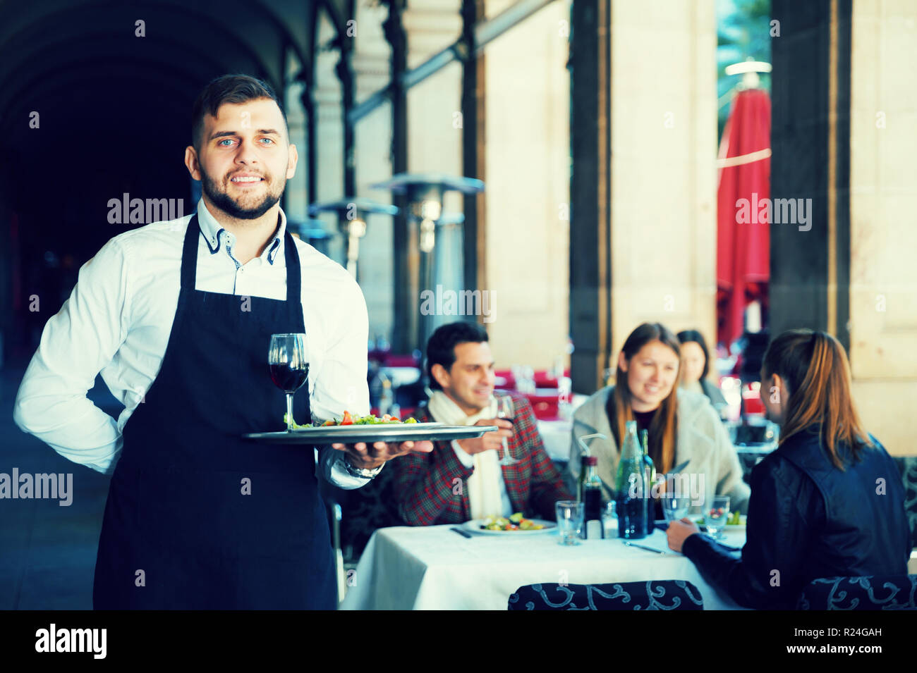 Cheerful waiter holding tray at restaurant with customers his behind ...