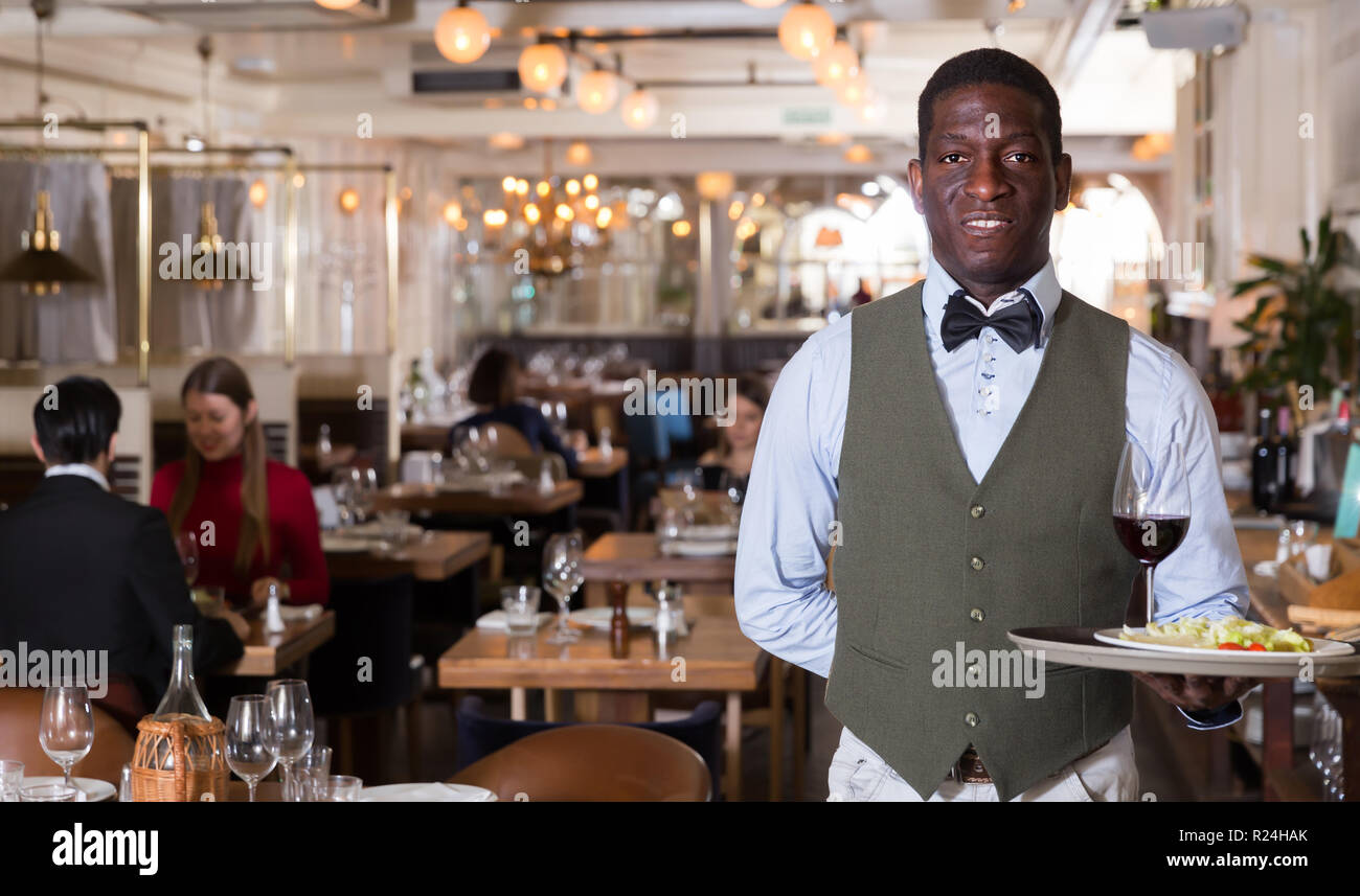 Young waiter who is standing with order in hall of luxurious restaurant ...