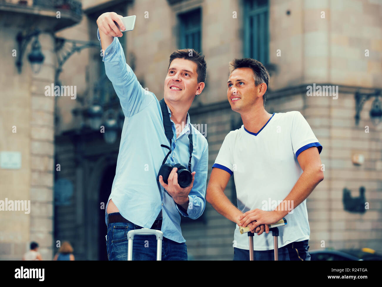 Two positive spanish men with luggage doing selfie during city tour at ...