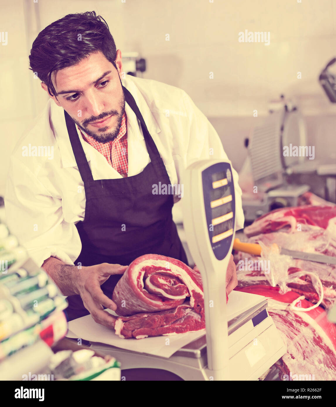 Adult male butcher in uniform is weighing meat for client in food shop ...