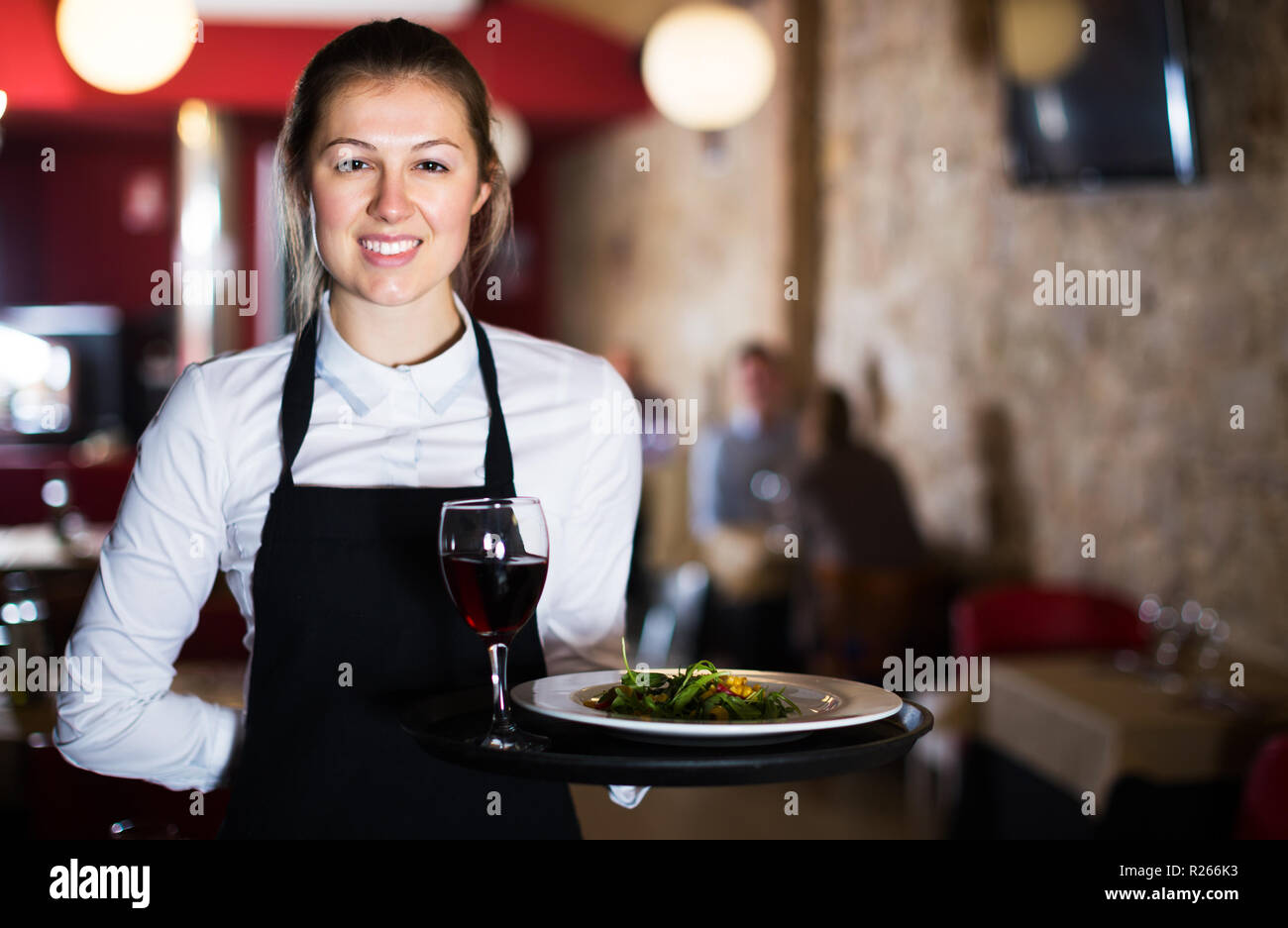Polite waitress wearing apron holding tray with dishes in restaurant ...