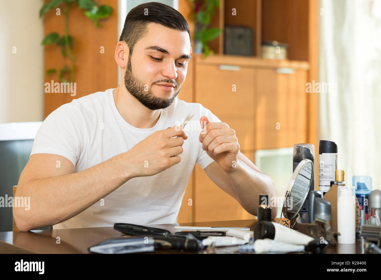 Handsome smiling young guy cutting nails with cuticle scissors in home ...