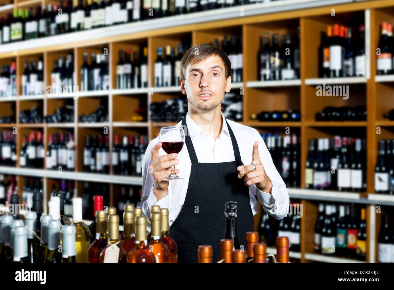 Happy young man seller wearing uniform holding glass of wine in wine ...