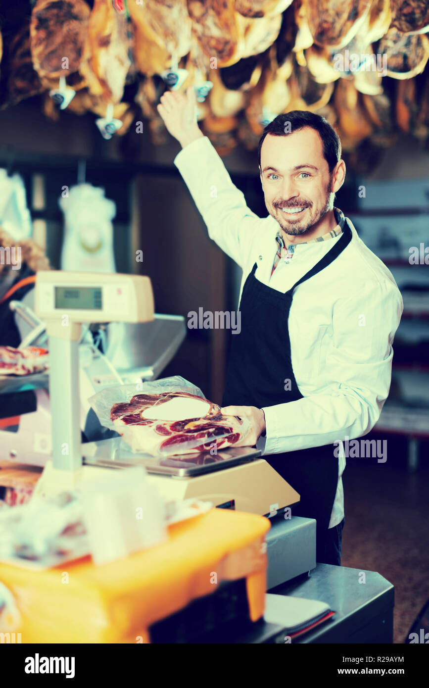 Smiling man seller weighing on scales piece of meat in butcher’s shop ...