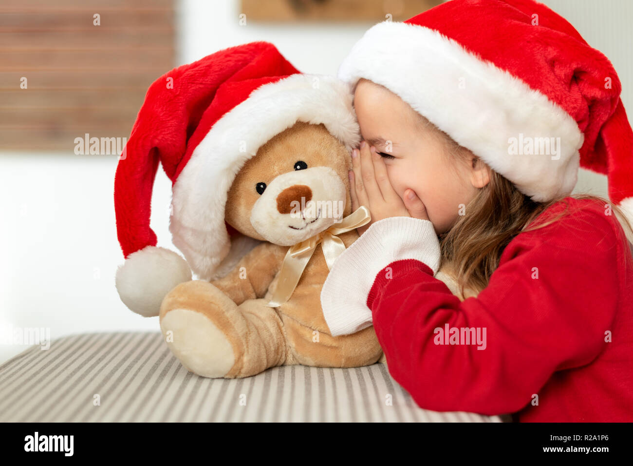 Cute young girl wearing santa hat whispering a secret to her teddy bear ...