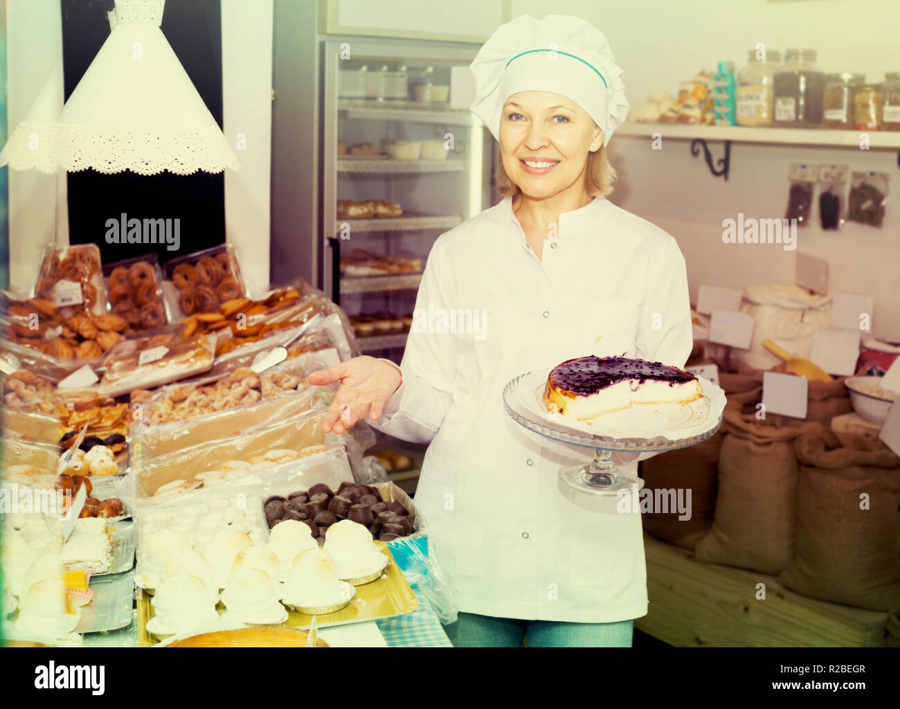 Portrait of positive mature female baker with pastry smiling in bakery ...