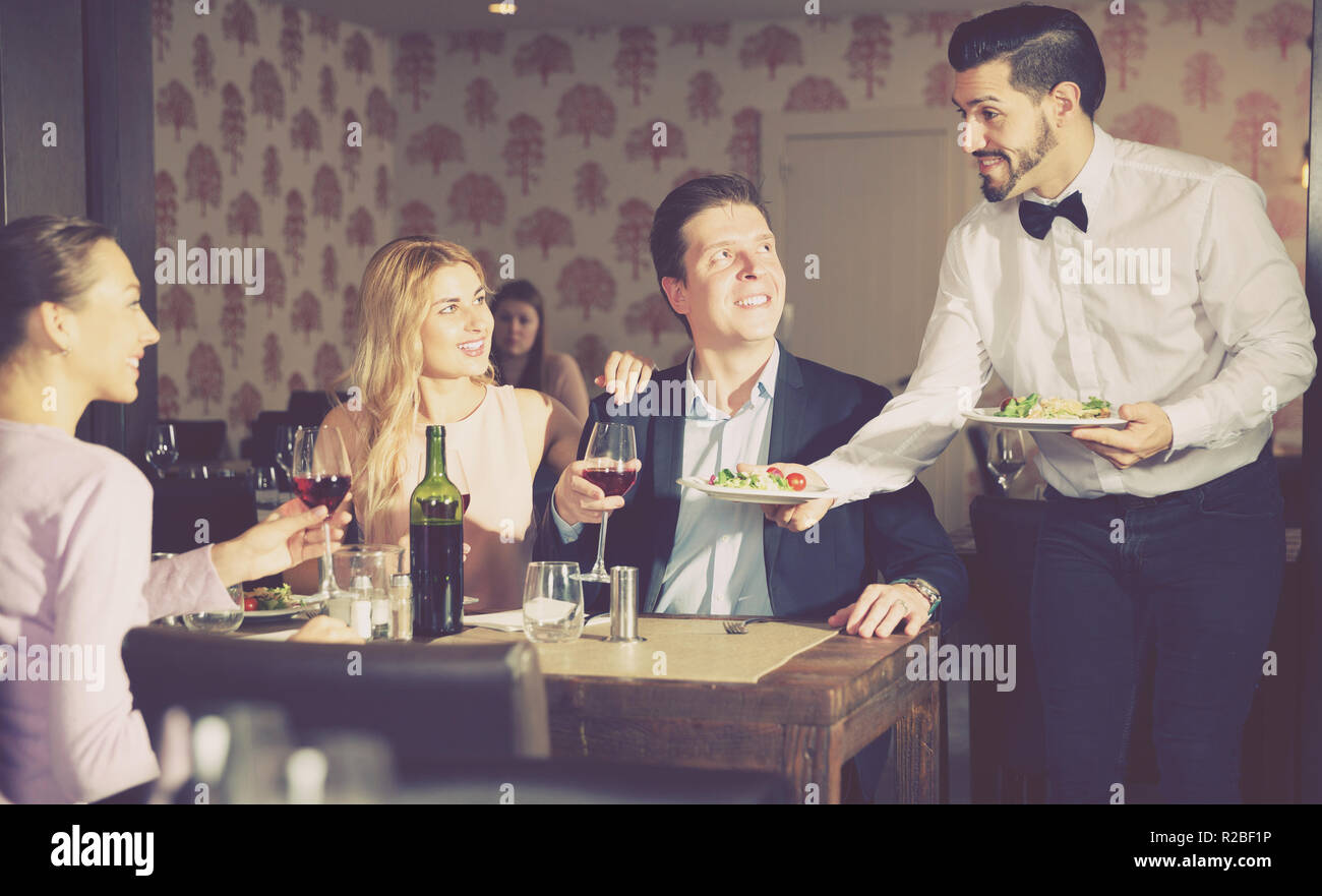 Polite adult waiter bringing ordered dishes to guests at restaurant ...
