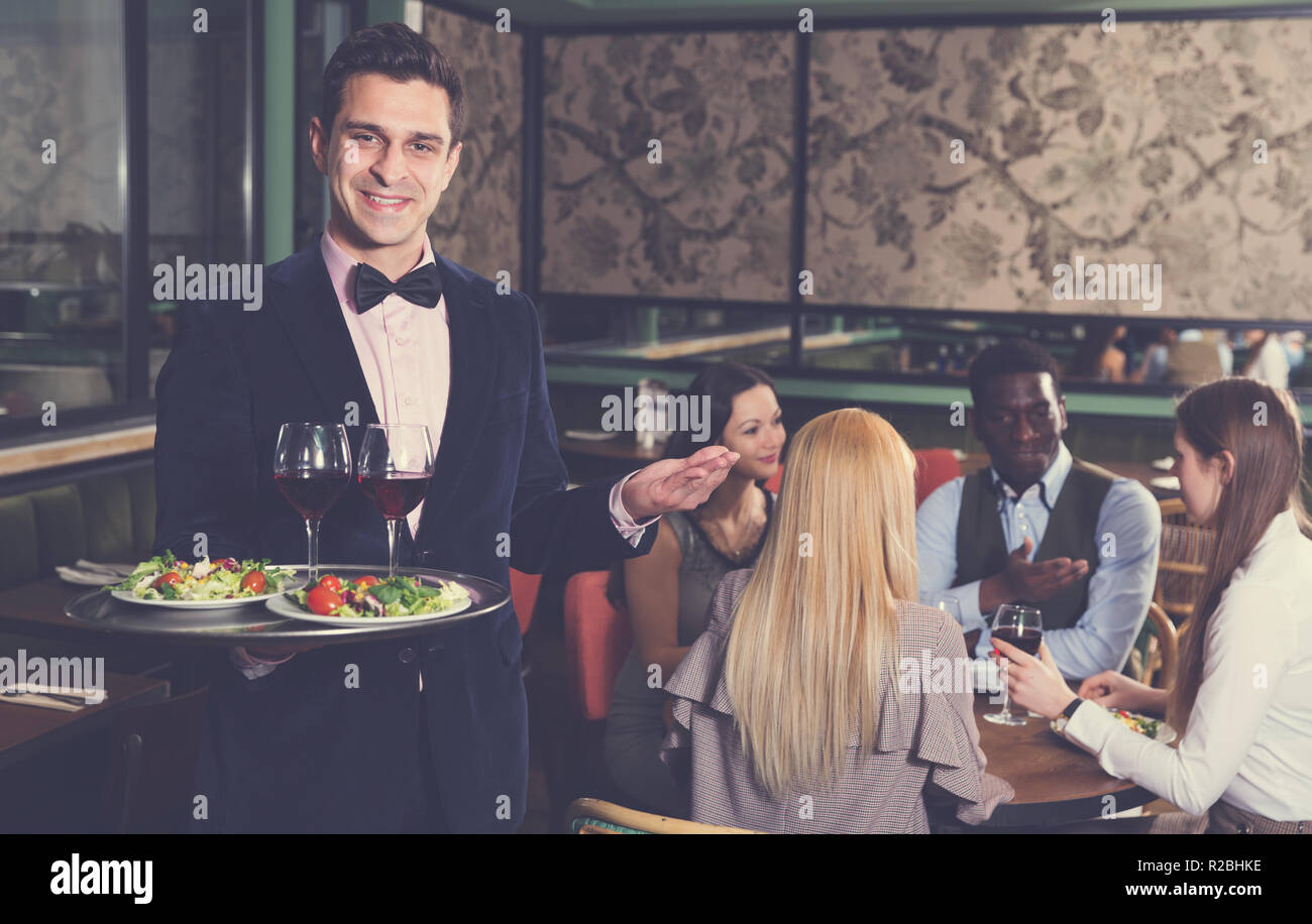 Polite waiter holding tray at restaurant with customers behind him ...